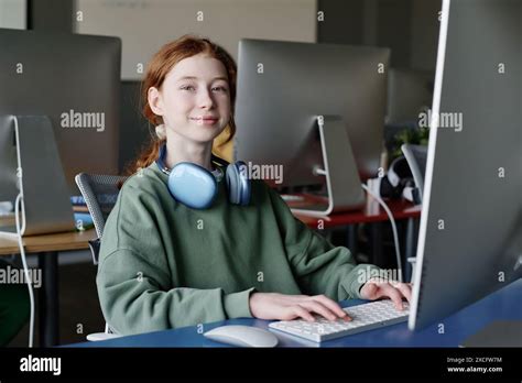 Medium Portrait Of Cheerful Teen Girl Wearing Headphones Sitting At Desk In Modern Classroom
