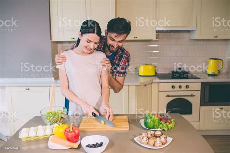 흥분 하 고 수염이 있는 사람은 여자 뒤에 서 서 음식을 본다 그는 어깨에 여자를 보유 하 고 있습니다 여자는 또한 아래로 보인다 그녀는 책상에 약간의 치즈를 잘라냅니다