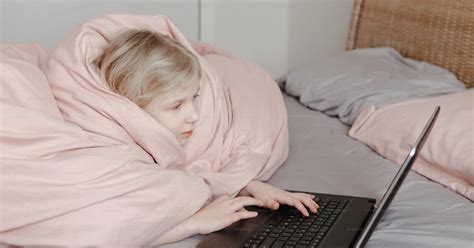 지루한 소녀는 담요 아래 침대에 누워 가상 온라인 학교 수업에서 학습합니다 집에서 노트북 인터넷으로 일하는 아이 무선 기술을