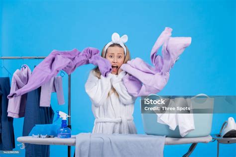한 소녀가 스튜디오의 화려한 파란색 배경에 서 있습니다 정렬 된 옷에 지루한 그녀는 다리미판 위에 옷을 던지며 그녀가 가운을
