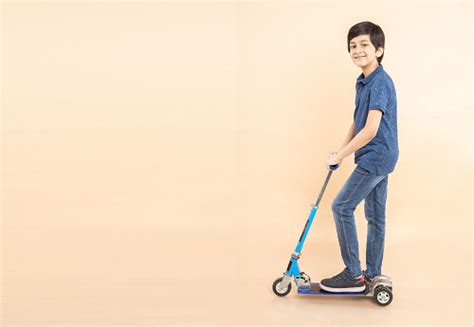 캐주얼한 옷을 입은 쾌활한 인도 소년은 복사 공간이 있는 베이지색 배경에 스쿠터를 탄다 스튜디오 촬영 어린 시절 및 활동적인