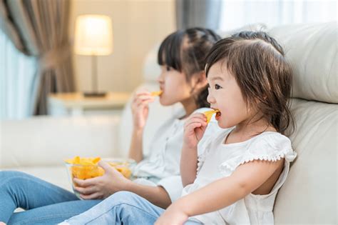 집에서 소파에 누워있는 아시아의 두 달콤한 자매 어린 소녀는 패스트 푸드 감자 칩을 먹는 것을 즐깁니다 배고픈 유치원 귀여운