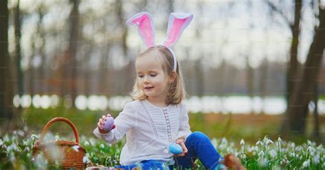 부활절에 달걀 사냥을 하는 토끼 귀를 입은 소녀 많은 헌병 꽃과 함께 잔디에 앉아 바구니에 다채로운 계란을 모으는 유아 야외에서 부활절을 축하하는 어린 아이 사진