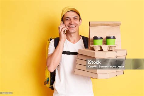 노란색 배경에 고립 된 흰색 티셔츠와 모자에 써모 배낭을 들고 매력적인 배달원을 웃고 고객과 전화하고 긍정적 인 감정을 표현하고
