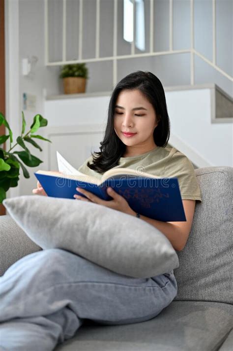 Beautiful Young Asian Woman Sits On The Comfy Sofa And Reads A Novel