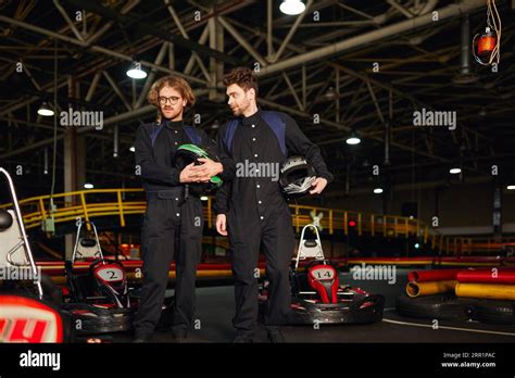 Two Kart Racers Standing Near Racing Cars And Holding Helmets Male