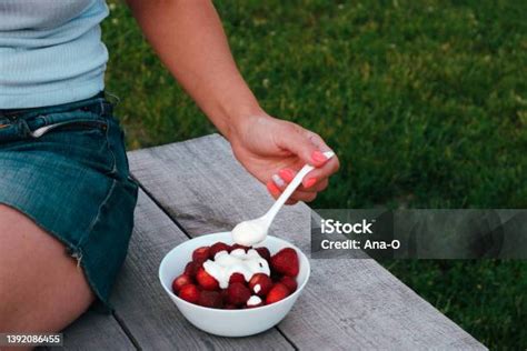 녹색 잔디밭에 나무 벤치에 앉아 데님 반바지를 입은 소녀가 숟가락으로 아침 식사를 위해 크림과 함께 딸기 디저트를 먹습니다 딸기가 있는 손과 흰색 접시의 클로즈업 보기