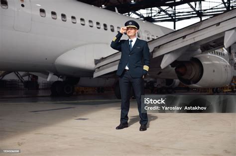 공항 격납고에서 큰 여객기 앞에 서서 모자를 조정하고 제복을 입은 자신감 있는 조종사의 전체 길이 샷 30 34세에 대한 스톡 사진 및 기타 이미지 Istock