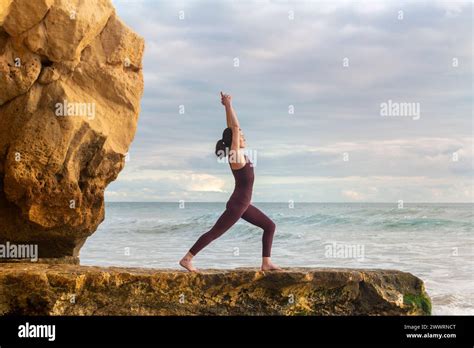 Woman Practicing Yoga Standing On A Rock By The Ocean Arms Above Head
