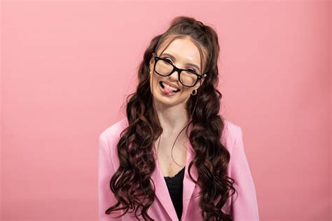 곱슬 머리로 혀를 내밀고 다니는 안경을 쓰고 멍청한 얼굴 농담 웃는 얼굴 핑크 스튜디오 배경에 괴상한 소녀를 가진 쾌활한 소녀