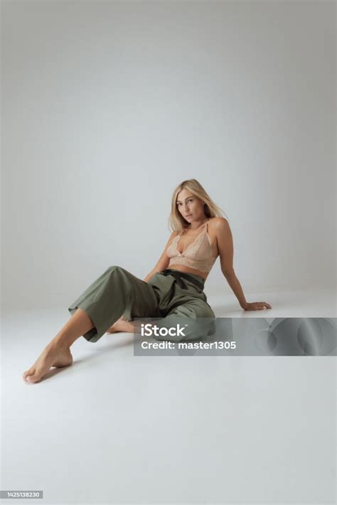긴 금발 머리와 자연스러운 메이크업을 한 아름다운 여성은 레이스 탑과 캐주얼 바지를 입고 회색 배경에 고립 된 바닥에 앉아 있습니다 30 34세에 대한 스톡 사진 및 기타