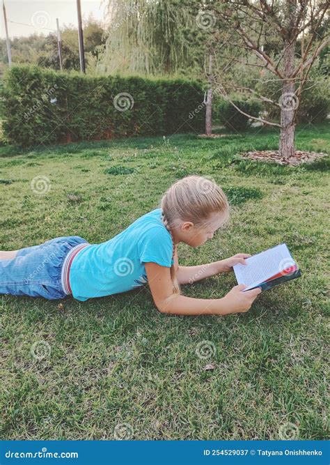 A Little Blonde Girl Is Lying On The Grass And Reading A Book Stock