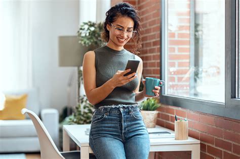 스마트 비즈니스 여성은 집에서 거실에서 테이블에 기대어 커피 한 잔을 마시면서 휴대 전화를 사용합니다 미소에 대한 스톡 사진 및