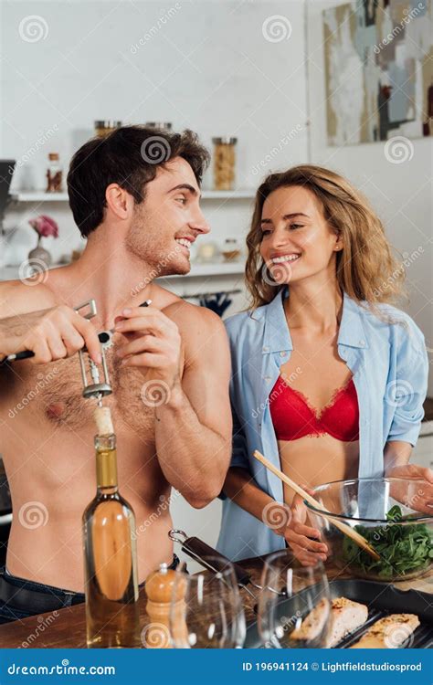 Shirtless Man Opening Bottle Of Wine And Looking At Girlfriend In