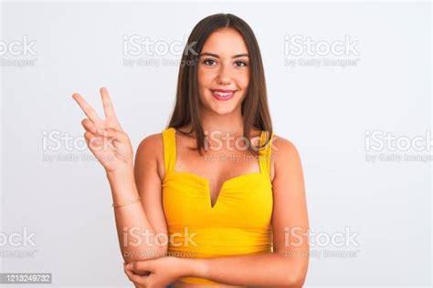 고립 된 흰색 배경 위에 서있는 노란색 캐주얼 티셔츠를 입은 젊은 아름다운 소녀는 승리의 표지판을하고있는 카메라에서 행복한 얼굴 윙크와 미소 2번 T 셔츠에 대한 스톡 사진