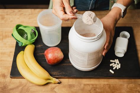청바지와 셔츠를 입은 여자는 하얀 캡슐 셰이커 바나나 사과가 있는 나무 테이블 위에 있는 플라스틱 병 위에 유청 단백질 분말의 일부가 담긴 계량 스푼을 들고 있다 단백질
