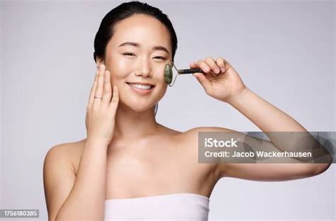 얼굴 미용 및 스킨 케어는 회색 배경의 스튜디오에서 얼굴에 롤러를 사용하는 모델 아시아 여성과 함께합니다 마사지 인물 사진 및 제품을 들고 있는 매력적인 젊은 여성과 함께