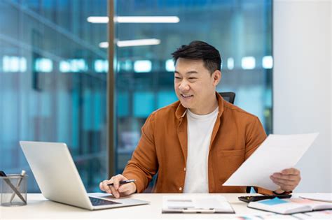 현대적인 사무실 환경에서 문서를 들고 노트북으로 작업하면서 미소 짓는 아시아 사업가는 전문적인 성공과 초점을 반영합니다 비즈니스 생산성 및 기술의 개념 경영자에 대한 스톡