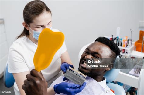 치과 치과 진료소에서 아프리카 환자의 치아 미백을 위한 치아 색조 가이드를 보여주는 치과 의사 아프리카는 의사의 손에 있는