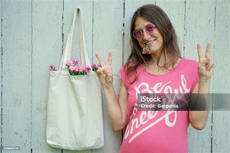 비명과 분홍색 둥근 안경을 쓴 분홍색 티셔츠를 입은 미소 짓는 히피 여성은 빈 캔버스 에코 쇼핑백 근처에 꽃이 들어와 평화 표지판을 만들었습니다 제로 폐기물 모형 35 39
