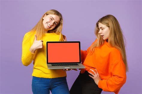 두 젊은 금발 쌍둥이 자매 여자는 파스텔 보라색 파란색 배경에 고립 된 빈 빈 화면으로 노트북 Pc 컴퓨터를 들고 엄지손가락을 보여주는 사람들 가족 라이프 스타일 개념입니다