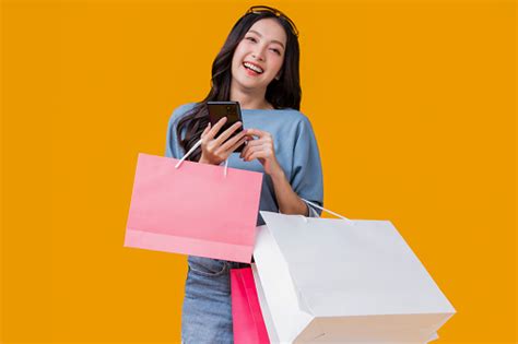 아시아 행복한 여성 여성 소녀는 노란색 배경 스튜디오 샷에 서있는 다채로운 쇼핑 패키지를 들고 있으며 가방이있는 카메라를보고