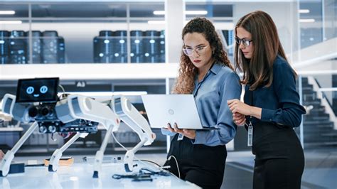 두 명의 산업용 로봇 공학 엔지니어가 로봇 모바일 프로토 타입으로 테이블에 모였습니다 두 명의 여성이 프로젝트에 대해 토론하고 랩톱 컴퓨터로 로봇 개를 프로그래밍합니다