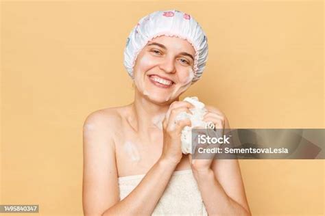 베이지색 배경 위에 고립된 샤워 모자를 쓴 예쁜 아름 다운 여자는 아침에 목욕을 한 후 신선함을 즐기는 스폰지를 들고 카메라를