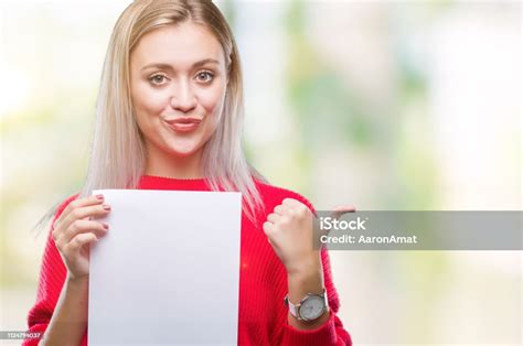 젊은 금발 여자 고립 된 배경 위에 빈 종이 시트를 들고 행복 한 얼굴 웃는 얼굴로 옆으로 엄지 손가락으로 표시 가리키기에 대한