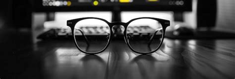 A Pair Of Blackframed Eyeglasses Rest On A Wooden Desk In Front Of A