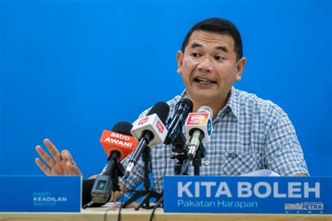 A Man Speaking Into Microphones In Front Of A Blue Wall With The Words