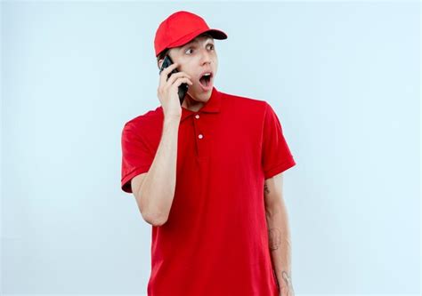 빨간 제복을 입은 젊은 배달 남자와 휴대 전화에 얘기하는 모자는 흰 벽 위에 서있는 충격을받은 식 무료 사진