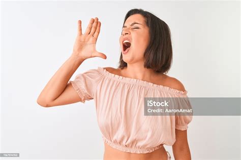 고립 된 흰색 배경 위에 서있는 캐주얼 한 티셔츠를 입은 젊은 아름다운 여성은 입에 손을 대고 큰 소리로 외치고 비명을 지르고