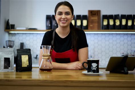 매력적인 긍정적인 젊은 여자의 반 길이 초상화에서 서 있는 빨간 앞치마를 입은 바와 카메라를 보고 세련 된 커피숍 인테리어에서 일 하는 검은 머리와 아름 다운 여성 바리