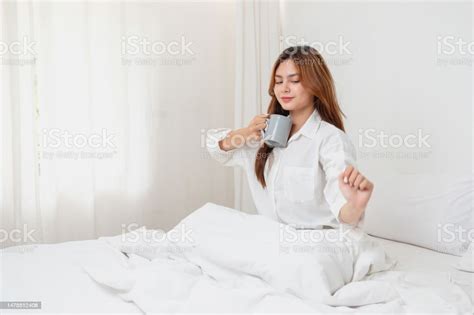 하얀 잠옷을 입은 여자가 주말 아침에 일어나 휴식을 취하고 노트북 휴대폰을 가지고 놀며 하얀 침실 안의 유리에서 빵을 먹고 차를