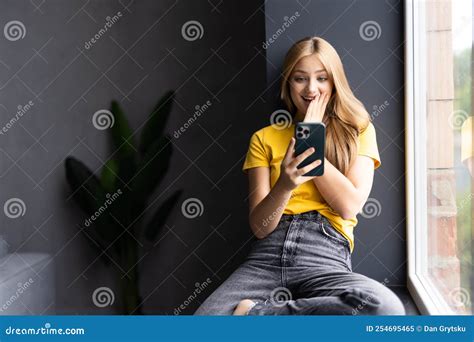 Attractive Young Woman Sitting On A Windowsill At Home And Texting On