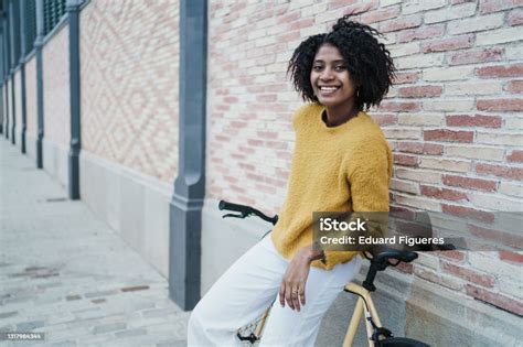 아프로 헤어 스타일 미소와 시원한 노란색 자전거와 분홍색 벽돌 벽 옆에 서 기뻐하는 평온한 흑인 여성 쿨한 태도에 대한 스톡