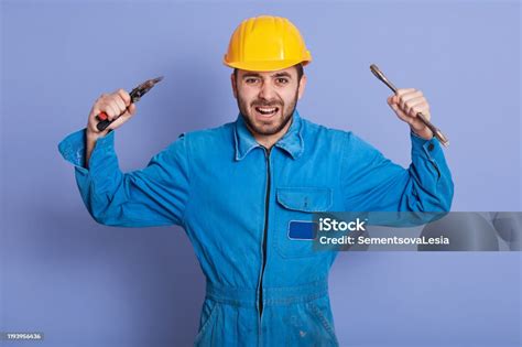 노란 헬멧과 파란색 유니폼을 입고 화가 난 건설 노동자 엔지니어의 초상화를 닫고 손을 들고 분노로 무언가를 외치고 수염이 있는