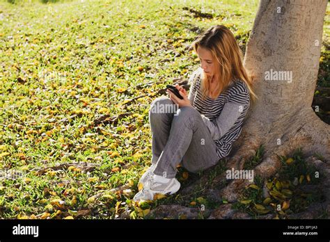 A Young Girl Sits Under A Tree At The Park Using Her Cell Phone Stock