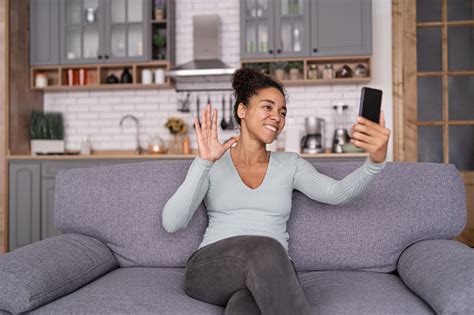 원격 통신 아프리카계 미국인 젊은 여성이 휴대전화에서 손바닥을 흔들며 부엌 방 내부의 소파에 앉아 있다 핸드폰으로 영상 통화를 하는 쾌활한 흑인 여성 집에 머물기 거리가