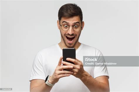 흰색 티셔츠를 입은 젊은 잘생긴 남자 둥근 안경을 쓰고 손에 전화를 들고 회색 배경에 고립 된 Omg 와우 얼굴로 화면을보고 전화에 대한 스톡 사진 및 기타 이미지 Istock