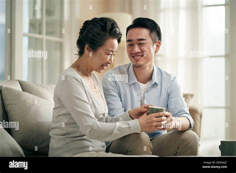 Asian Adult Son Sitting On Couch At Home Chatting With Senior Mother Happy And Smiling Stock