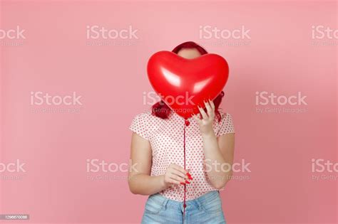 빨간 머리를 가진 여자는 분홍색 배경에 고립 된 그녀의 손에 큰 비행 빨간 하트 모양의 풍선 뒤에 그녀의 얼굴을 숨깁니다 20 24세에 대한 스톡 사진 및 기타 이미지