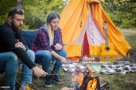 아름 다운 커플 앉아 캠프 파이어 주위에 캠핑 텐트와 배경에 남자 막대기로 화재 제스처 2명에 대한 스톡 사진 및 기타 이미지 Istock