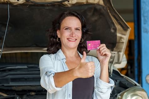 분홍색 신용 카드를 들고 엄지 손가락을 치켜 올리는 중년 백인 여성 자동차 차고의 열린 자동차 후드 앞에 서서 성공적인 자동차 수리 서비스에 대한 승인과 자신감을 보여줍니다