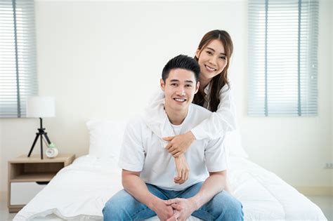 아시아 의 새로운 결혼 부부의 초상화는 침대에 앉아 카메라를 보고 파자마에 매력적인 아름다운 젊은 남자와 여자는 집에서 침실에서