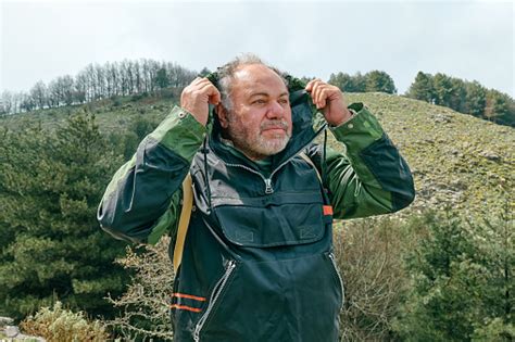 성숙한 수염을 기른 여행자 남자는 산림에서 하이킹을 하고 머리에서 재킷의 후드를 벗고 계곡에서 신선한 산 공기를 즐깁니다 60 64세에 대한 스톡 사진 및 기타 이미지