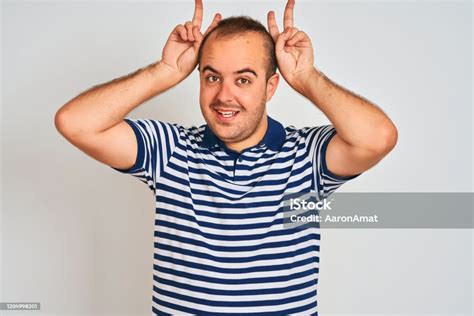 고립 된 흰색 배경 위에 서있는 캐주얼 스트라이프 폴로를 입은 젊은 남자 토끼 귀로 머리에 손가락으로 재미있고 미친 포즈를 취하고 쾌활한 미소를 지었습니다 귀 부분에 대한