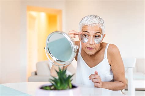 붓기 주름 다크 서클을 위한 아이 마스크 아래 눈 패치 개념 흰색 셔츠를 입은 수석 회색 머리 의 여성 거울을 보면서 실버 눈 패치를 적용 거울에 대한 스톡 사진 및 기타