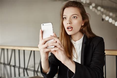 Удивленная молодая женщина смотрит на мобильный телефон сидя в кафе